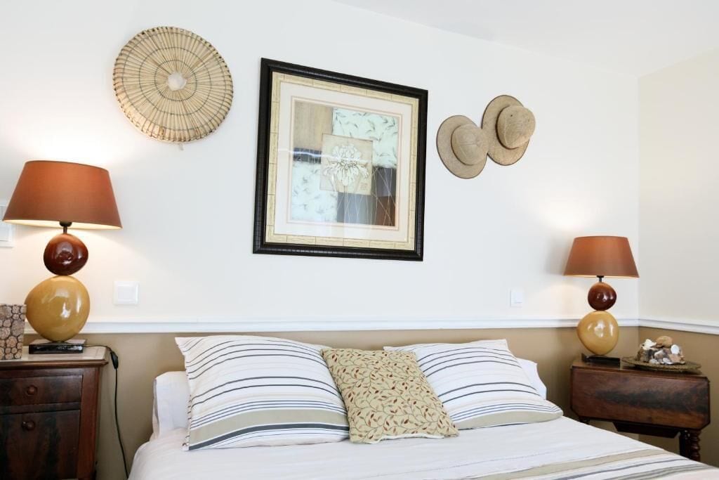 Quarto bem arrumado com uma cama, duas mesas de cabeceira com abajures, uma obra de arte emoldurada e elementos decorativos na parede.