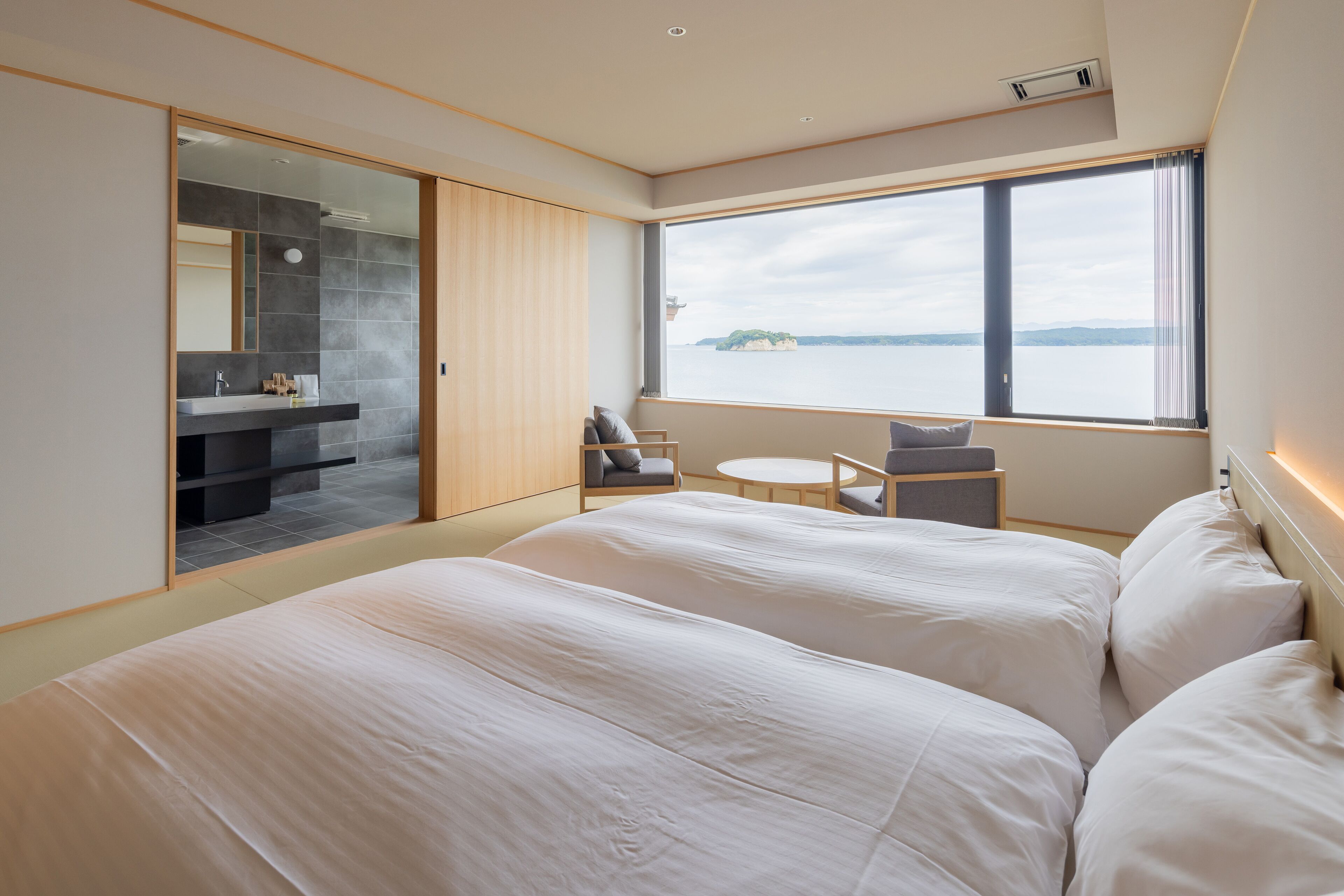 Quarto moderno com duas camas, uma mesinha redonda e vista para a água através de uma grande janela.