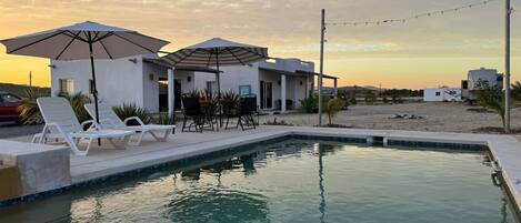 Piscina com espreguiçadeiras e guarda-sóis, um edifício branco e uma praia ao fundo.