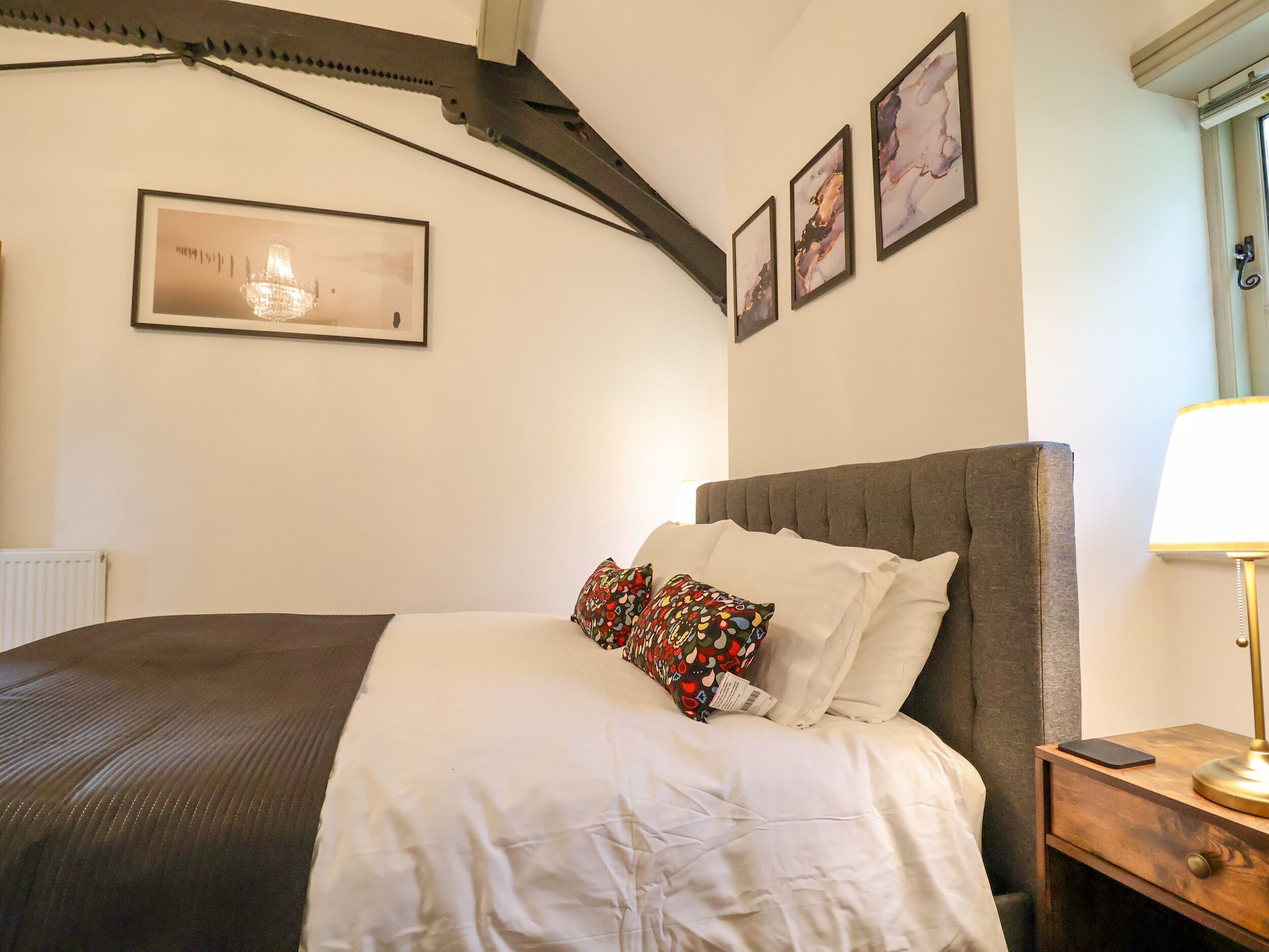 Cama bem arrumada com lençóis brancos e almofadas coloridas, mesa de cabeceira de madeira com abajur, e três quadros na parede.