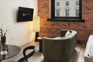 Quarto de hotel moderno com parede de tijolos, TV de tela plana, mesa redonda, cadeira, abajur e janela com vista para edifícios.