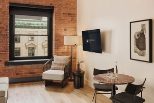 Quarto de hotel com parede de tijolos, uma janela grande, cama, poltrona, mesa com cadeiras, televisão e obras de arte.