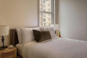 Quarto de hotel com cama, duas luminárias de cabeceira, mesa de cabeceira com telefone e xícara, e janela com vista para edifícios.