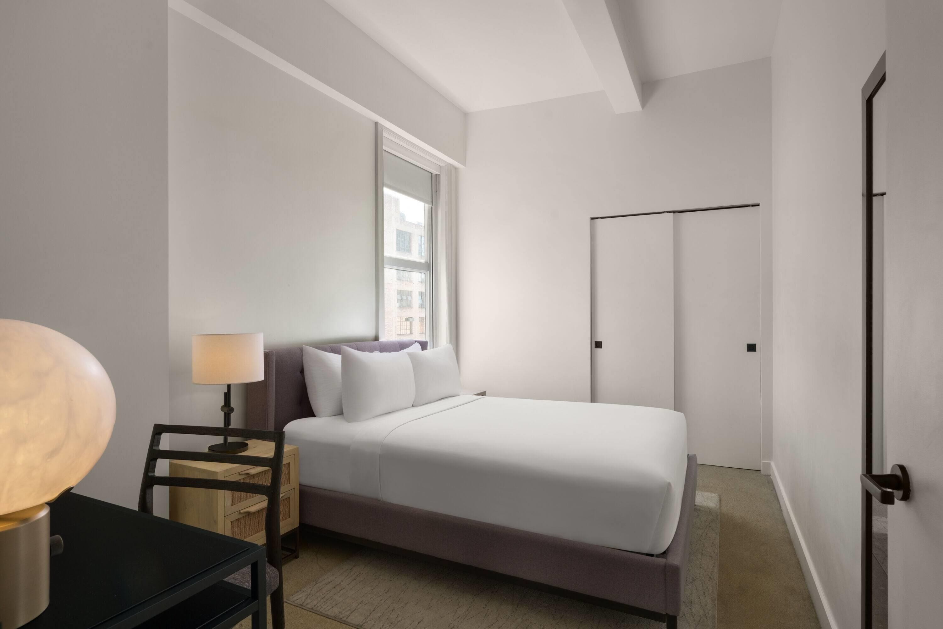 Quarto de hotel moderno com uma cama, uma escrivaninha com luminária, uma janela e uma porta.