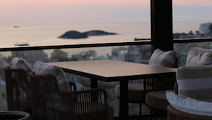Uma área de refeições com cadeiras de vime e uma mesa, com vista para um corpo d'água com barcos e terra ao longe.