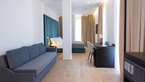 Quarto de hotel moderno com um sofá cinzento, uma cama com cabeceira azul-turquesa, uma mesa de escritório com uma cadeira e uma TV sobre um suporte preto.
