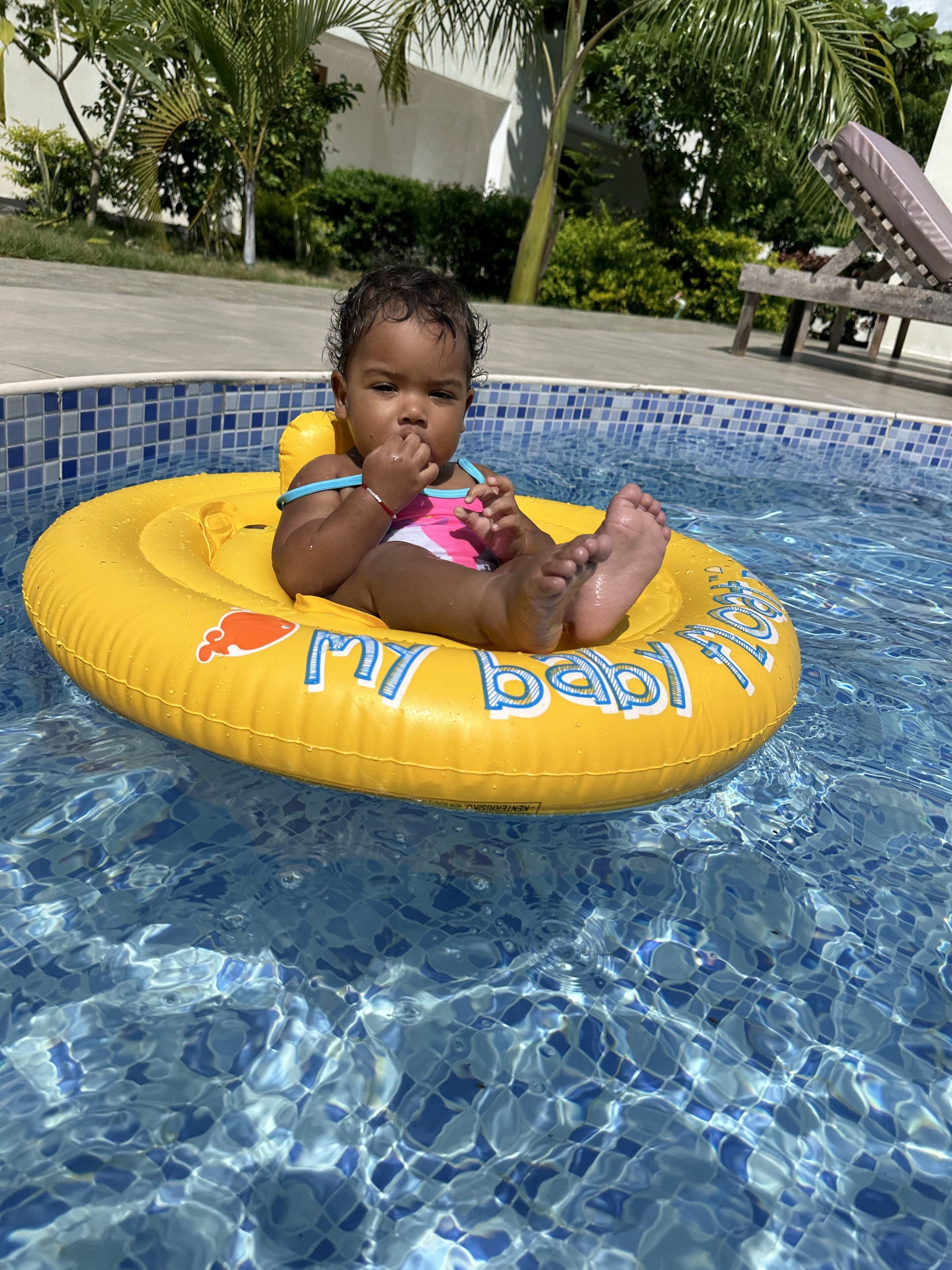 Criança em piscina inflável amarela com a inscrição 'Baby Beach', rodeada por água azul cristalina e borda de piscina azulejada.