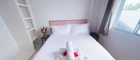 Cama bem arrumada com uma toalha branca enrolada e flores vermelhas, mesa de cabeceira com um vaso de flores, e janela com cortinas.