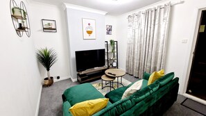 Uma sala de estar com um sofá verde, uma televisão, uma mesinha e um vaso de planta.