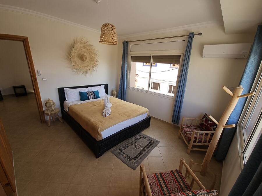 Quarto com cama, janela com cortinas, aquecedor, cadeira e mesa pequena com abajur.