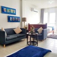 Quarto de hotel moderno com um sofá azul, uma poltrona menor, uma mesa redonda com um vaso de flores, uma cama com cabeceira de madeira e obras de arte na parede.