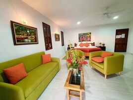 Quarto de hotel com sofá verde, mesa de centro de madeira com um vaso e cama com almofadas laranja.