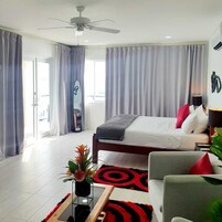 Quarto de hotel com cama, escrivaninha, poltrona, planta e ventoinha no teto.