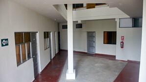 Um corredor com paredes brancas, chão vermelho e um pilar branco. Há uma porta com uma placa que diz 'SALIDA' e um extintor de incêndio na parede.