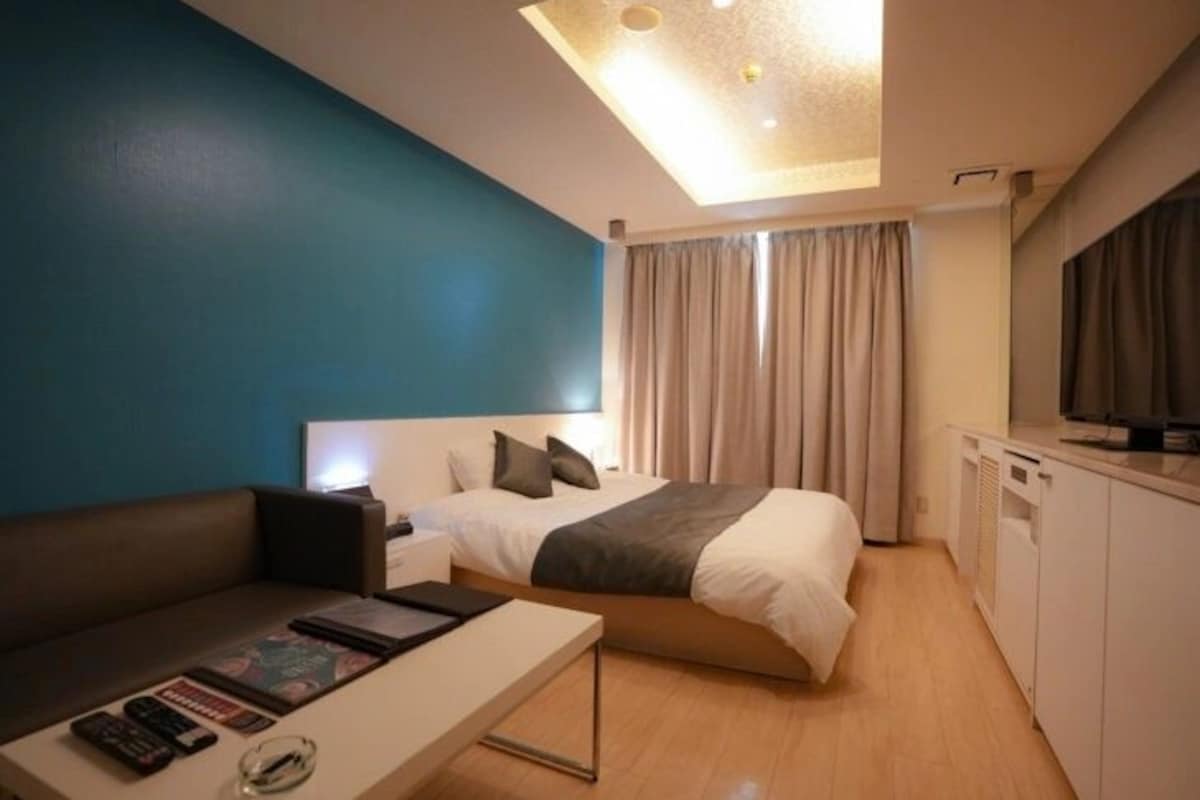 Quarto de hotel moderno com cama, escrivaninha com cadeira, televisão e área de cozinha compacta.
