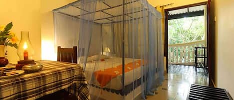 Quarto com uma cama sob um mosquiteiro, uma mesa com toalha xadrez e vista para o verde lá fora.
