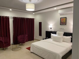 Quarto de hotel com cama, mesa de cabeceira, luminária, cadeira e cortinas.