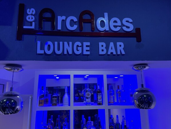 Um bar chamado "Les Arcades Lounge Bar", com prateleiras cheias de garrafas de bebidas e luzes de discoteca.