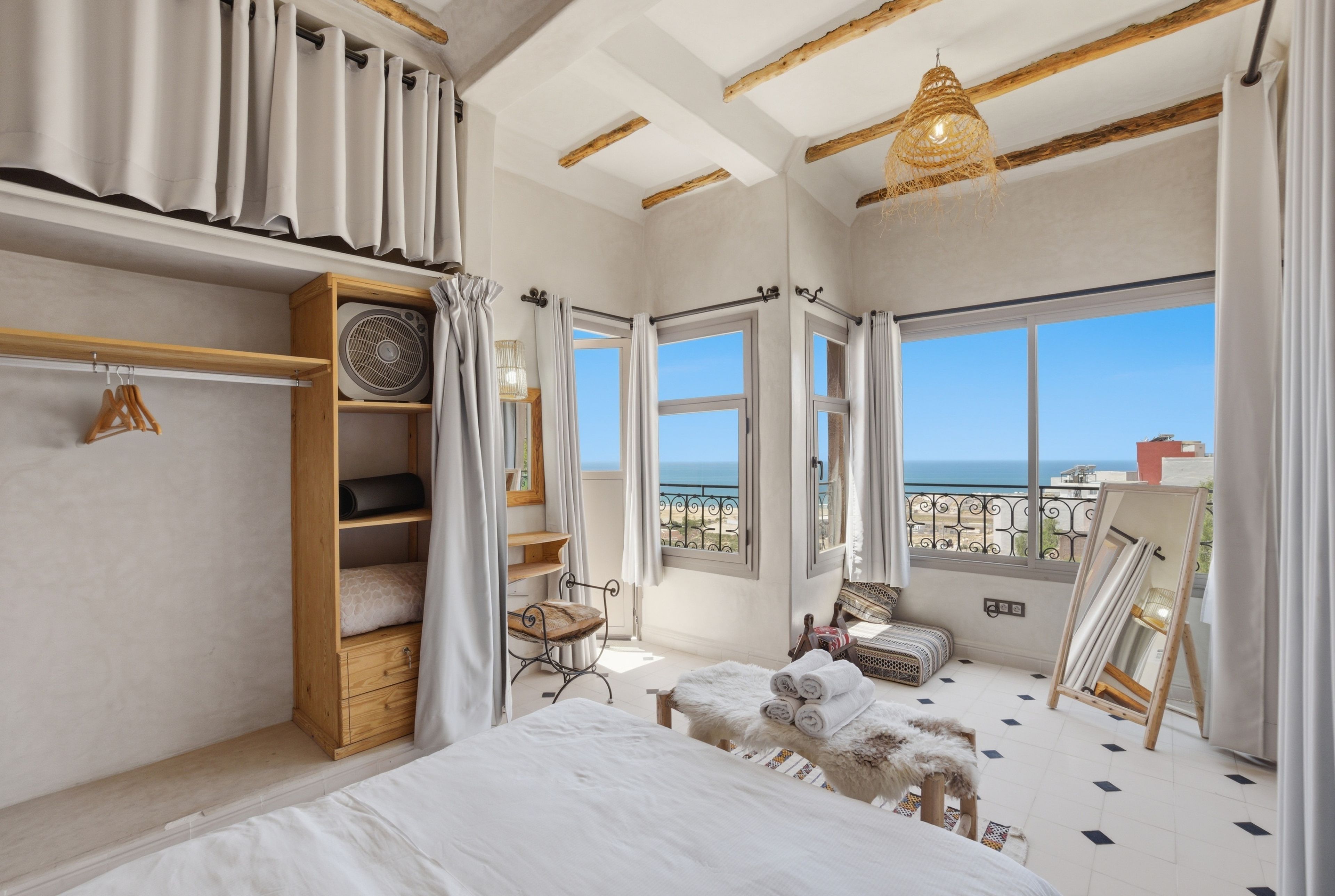 Quarto com cama, guarda-roupa de madeira, cadeira, mesa pequena e uma janela grande com vista para o mar.