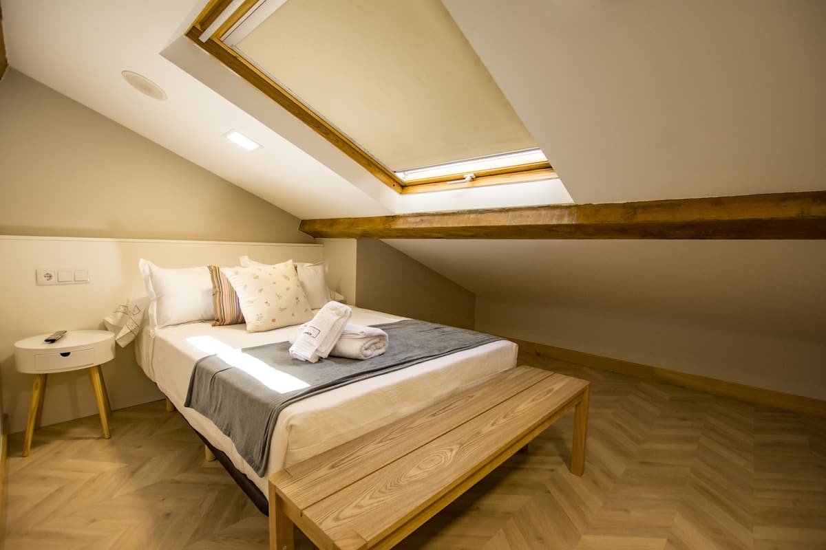 Quarto com cama, mesa de cabeceira e claraboia.