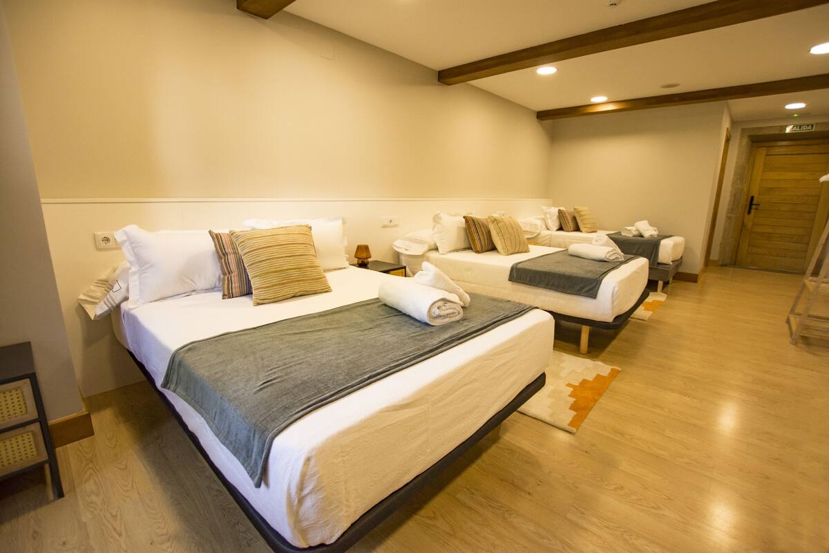 Quarto de hotel com duas camas, cada uma com manta e almofadas cinzas, uma pequena mesa de cabeceira com abajur e piso de madeira.