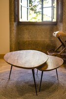 Uma mesa redonda de madeira com superfície texturizada e quatro pernas, colocada sobre um tapete circular em um quarto com parede de pedra e janela com grades.