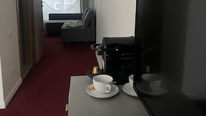 Quarto de hotel com televisão, máquina de café e uma chávena sobre uma mesa. Há uma janela com cortinas e vista para edifícios lá fora.