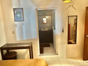 Quarto pequeno com cama, mesa, espelho e porta para um banheiro.