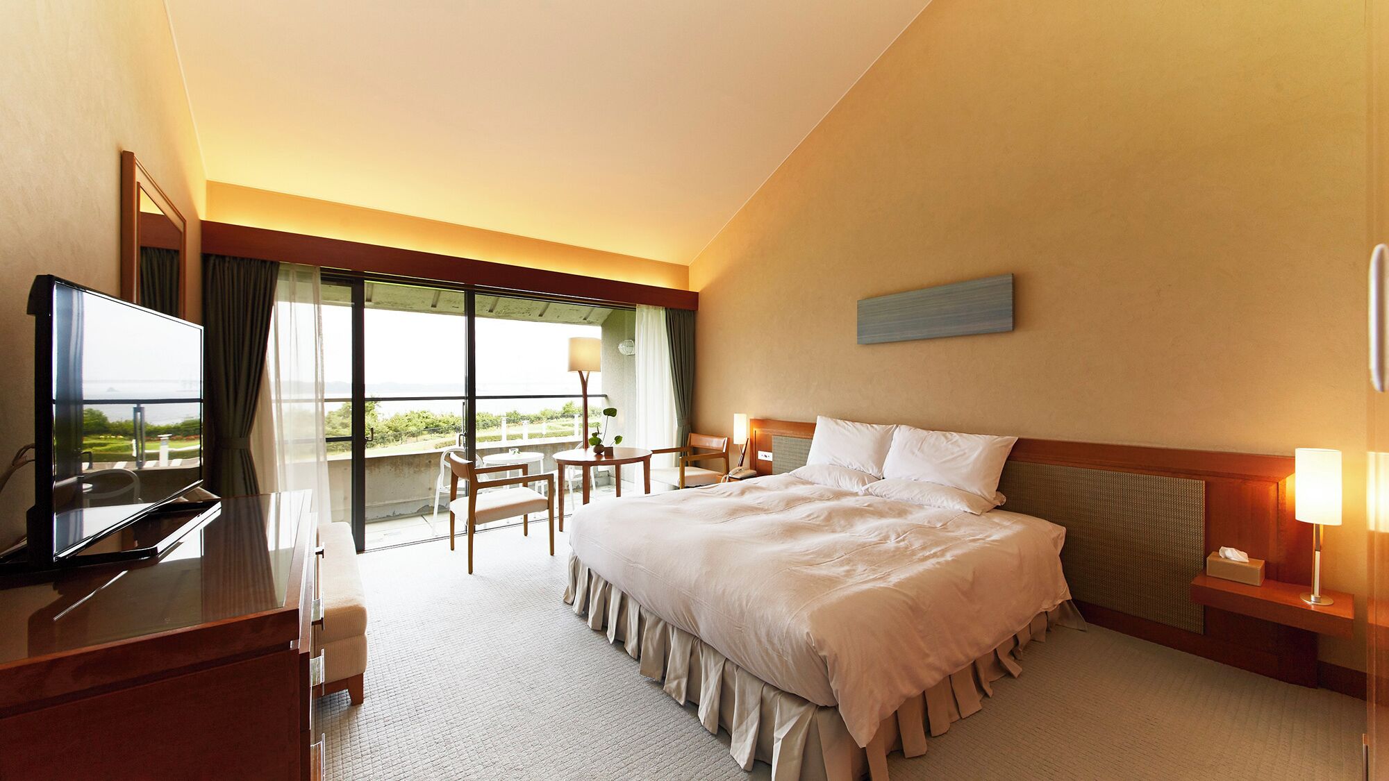 Quarto de hotel com cama, escrivaninha, cadeira, televisão, varanda com vista e mesa de cabeceira com abajur.