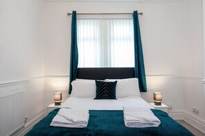 Cama bem arrumada com lençóis brancos, cortinas verde-água e cabeceira preta. Duas luminárias de cabeceira em cada lado da cama.
