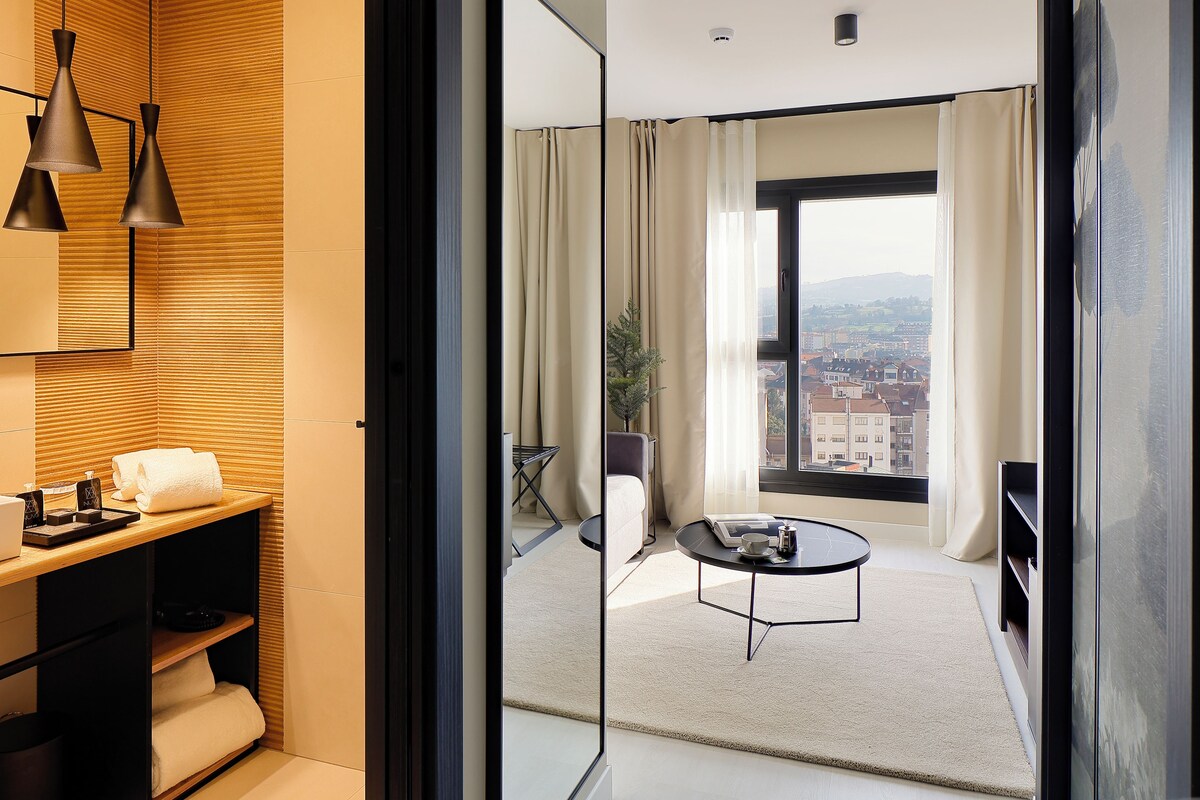 Quarto de hotel moderno com varanda, uma mesa de centro redonda e vista para a paisagem urbana.