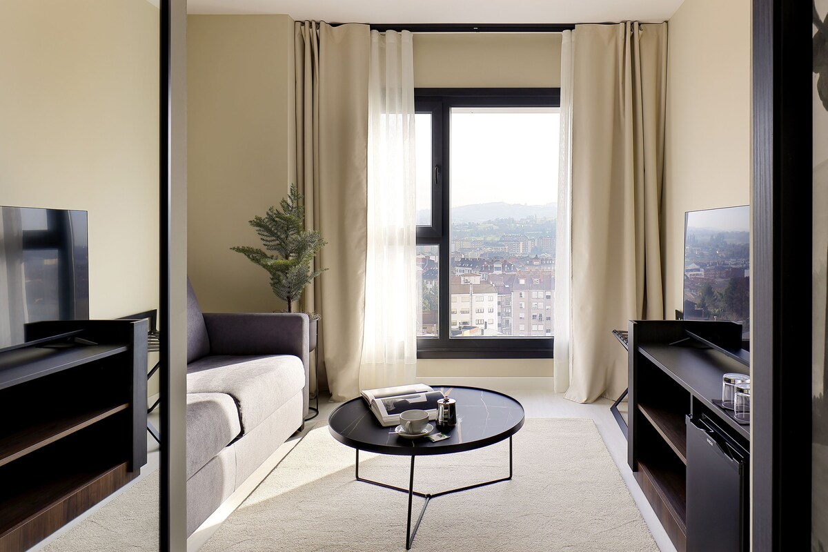 Quarto de hotel moderno com sofá, mesa de centro, TV e uma grande janela com vista para a cidade.