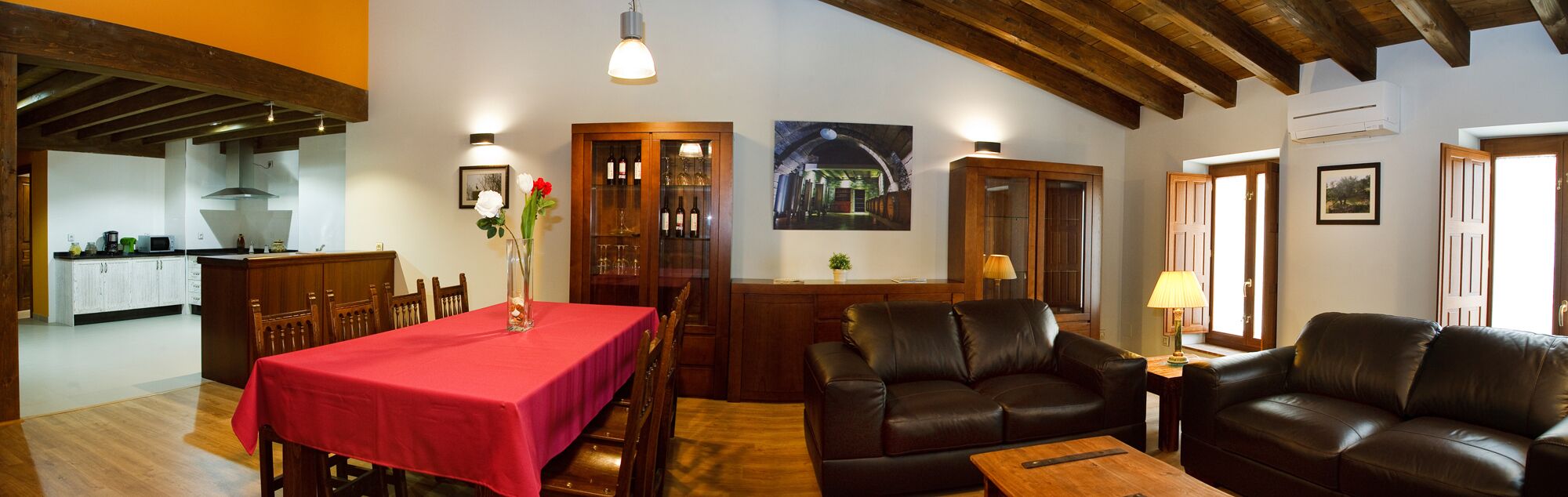 Uma área de refeições com toalha de mesa vermelha, um armário de madeira e uma mesa de jantar. Há um sofá de couro preto e uma mesa de centro de madeira.