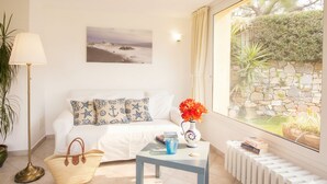Um quarto aconchegante com um sofá branco, uma mesa pequena com um vaso de flores, uma luminária e vista para um jardim pela janela.