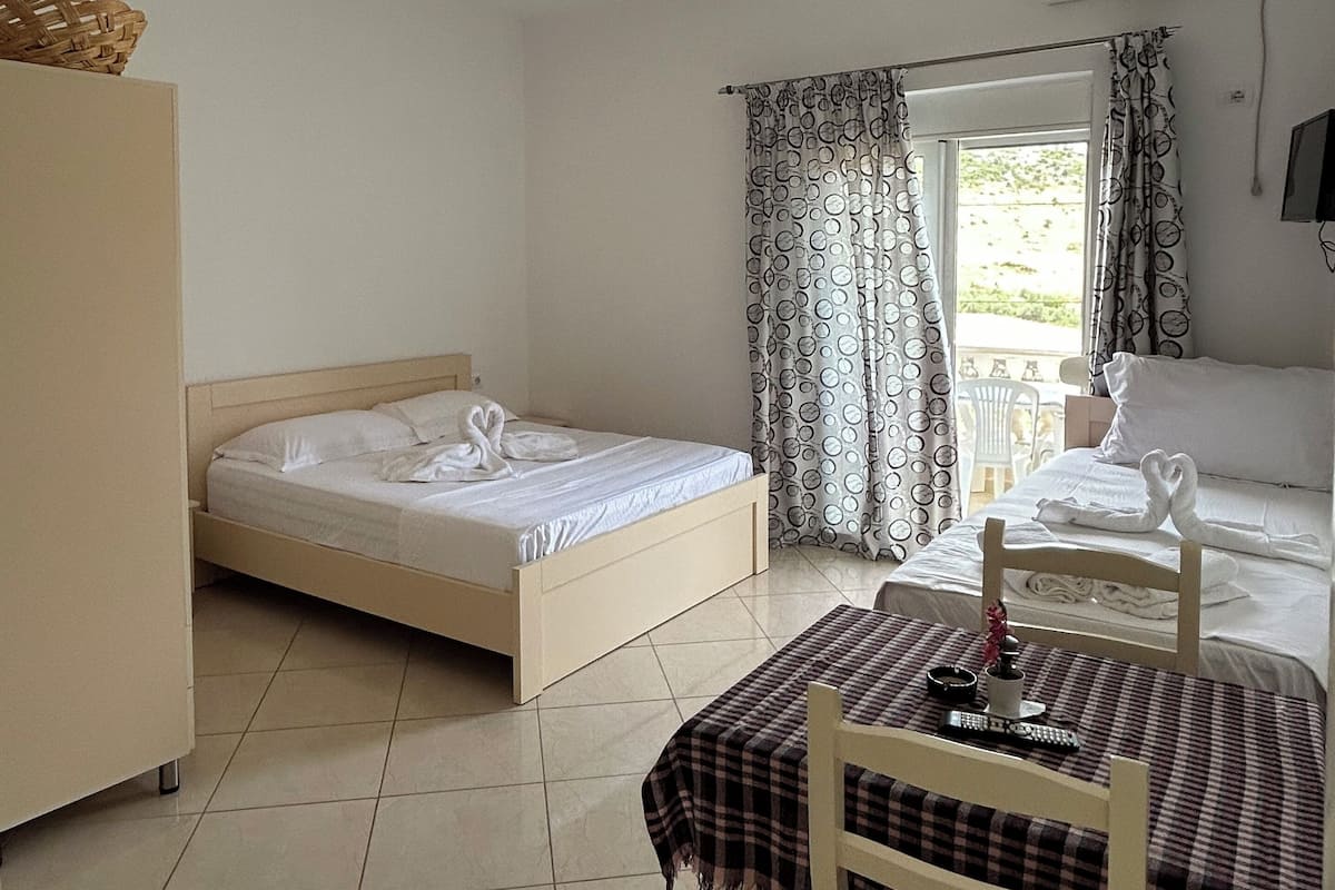 Quarto pequeno e limpo, com duas camas de solteiro, uma mesa de jantar com toalha xadrez, uma cadeira e uma janela com cortinas estampadas.