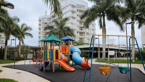 Um parquinho com escorrega e balan ços, cercado por palmeiras e um edifício ao fundo.