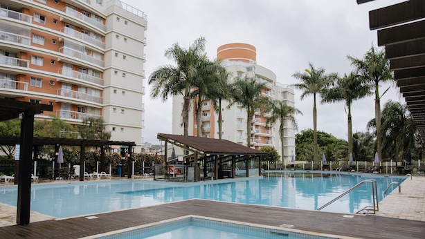 Piscina com deck de madeira, cercada por palmeiras e um edifício com várias andares e varandas.