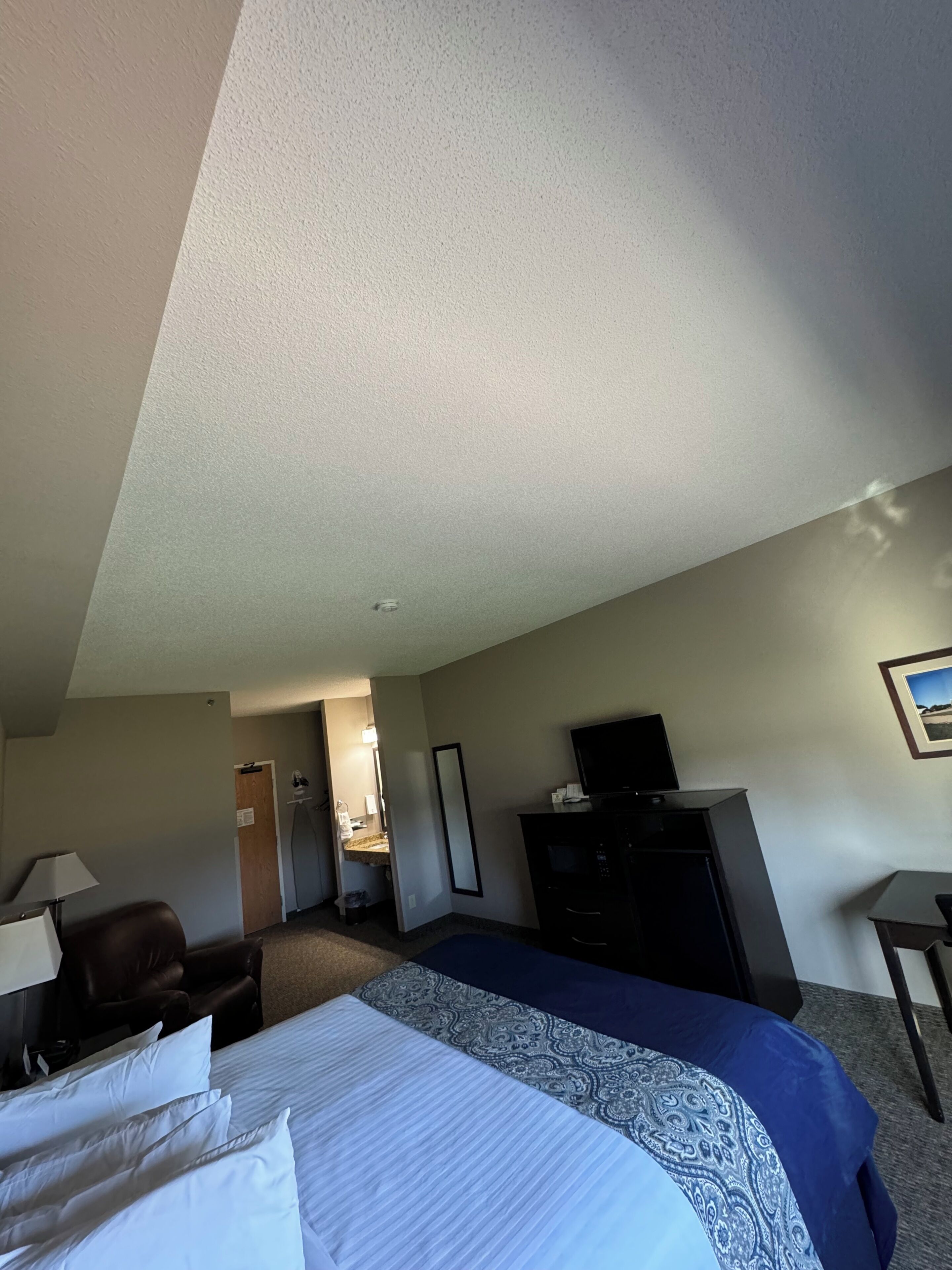 Quarto de hotel com cama, escrivaninha, cadeira, televisão e espelho.