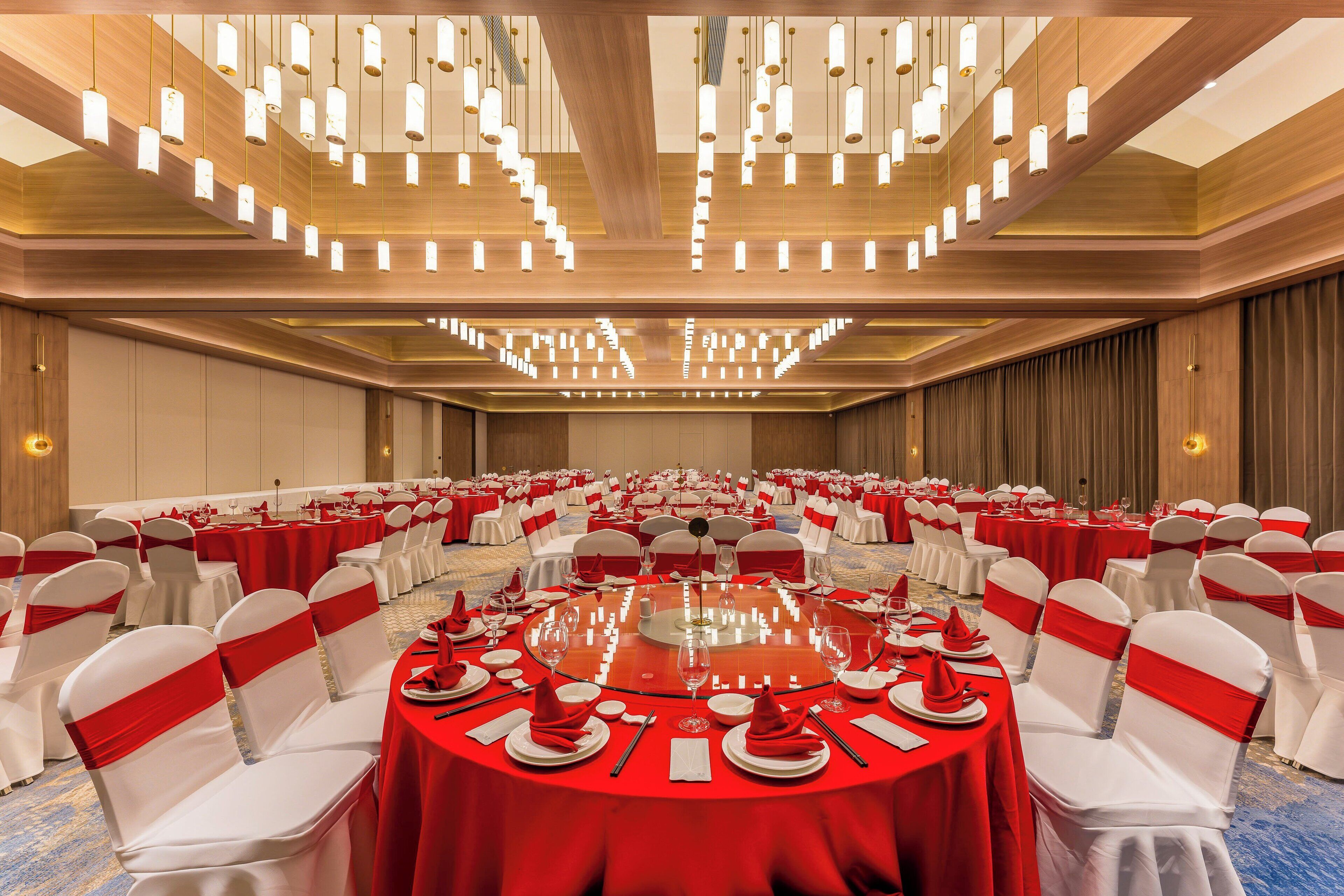 Salão de banquetes com mesas redondas, cadeiras brancas e fitas vermelhas, e um lustre central.