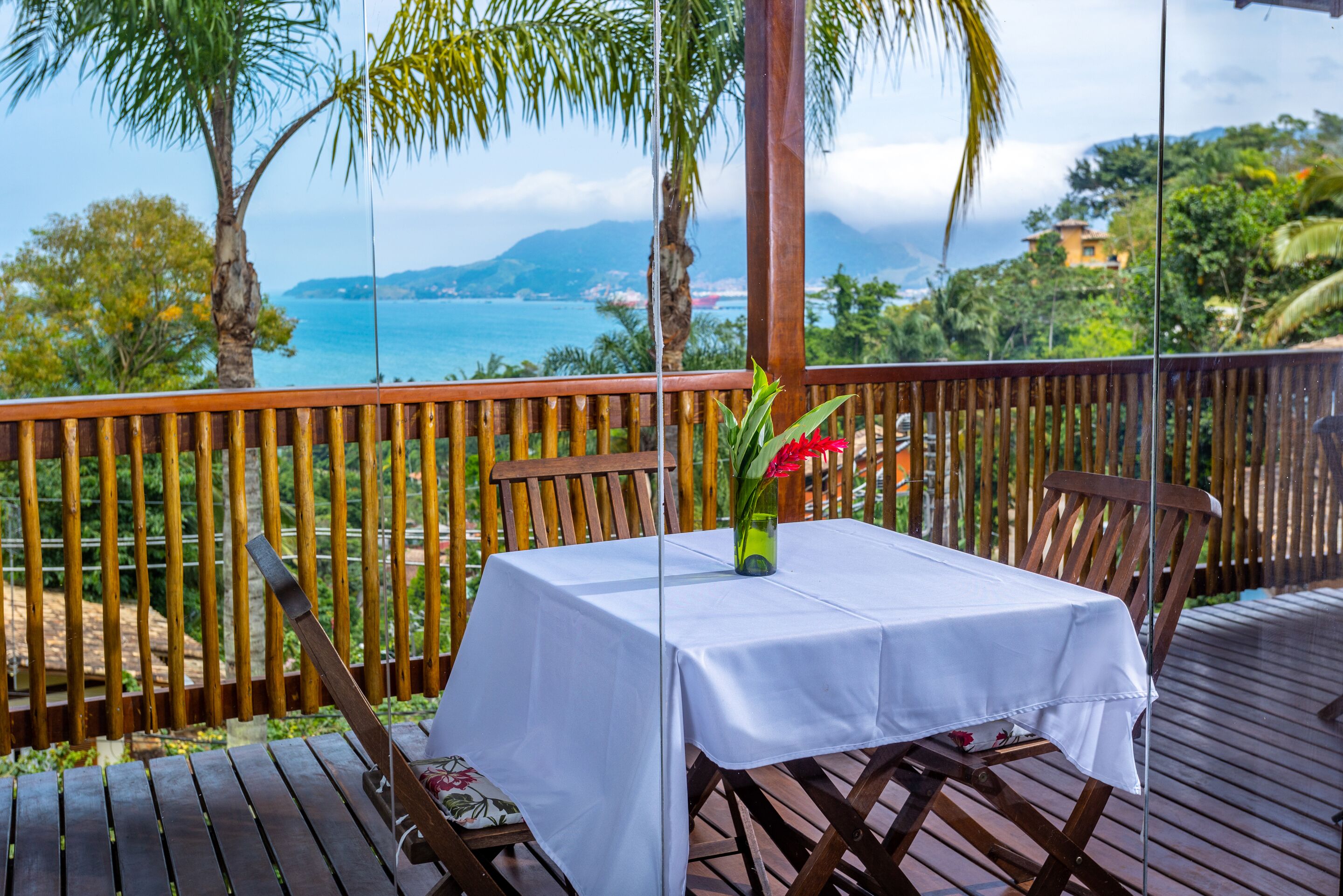 Um deck de madeira com uma mesa posta para dois, com vista para uma paisagem costeira com montanhas e um corpo de água.