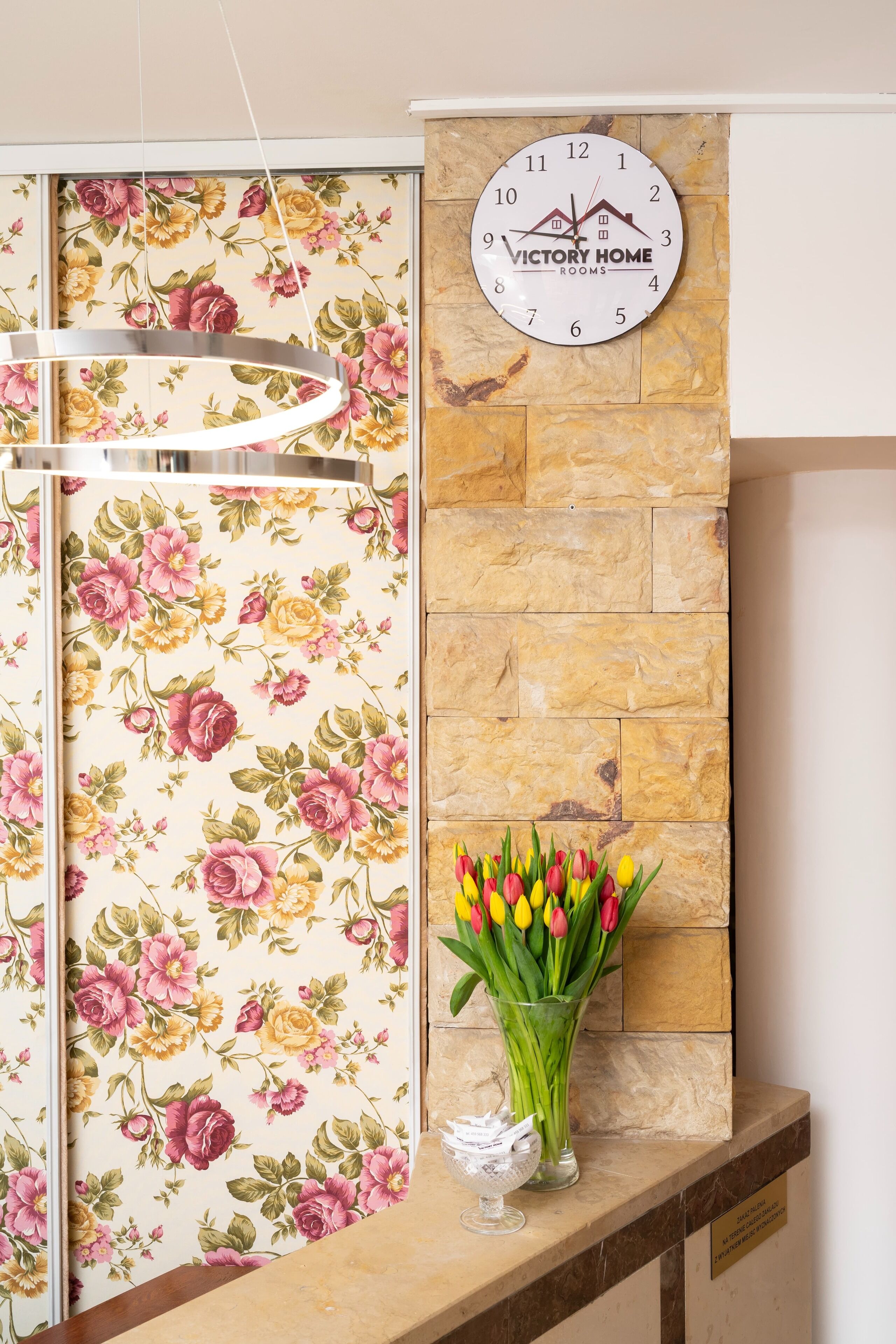 Uma área de recepção com papel de parede floral, um relógio e um vaso com tulipas.