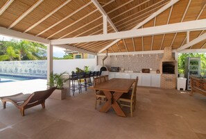 Um pátio externo com mesa e cadeiras de madeira, um forno de tijolos e uma piscina ao fundo.