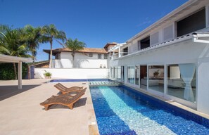 Uma piscina com espreguiçadeiras, um edifício branco com grandes janelas e palmeiras.