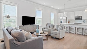 Uma sala de estar moderna com um sofá cinzento, poltronas e uma mesa de centro. Uma televisão de ecrã plano está montada na parede. A área da cozinha inclui armários brancos, um micro-ondas e um frigorífico.