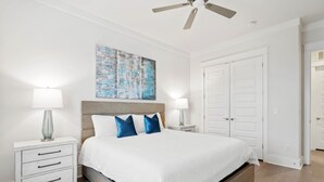 Quarto com cama, duas mesas de cabeceira, ventoinha no teto, uma grande obra de arte e um guarda-roupa.