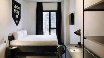 Quarto de hotel com cama, escrivaninha, cadeira, luminária de mesa, televisão e janela com cortinas.
