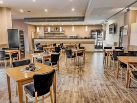 Um café moderno com mesas de madeira e cadeiras pretas, uma área de bar com prateleiras cheias de garrafas e um chão de madeira em padrão herringbone.