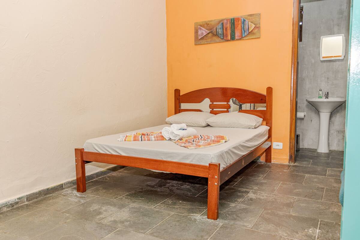 Uma cama de solteiro com estrutura de madeira e colchão branco, uma pequena mesa de cabeceira e um quadro pendurado na parede.