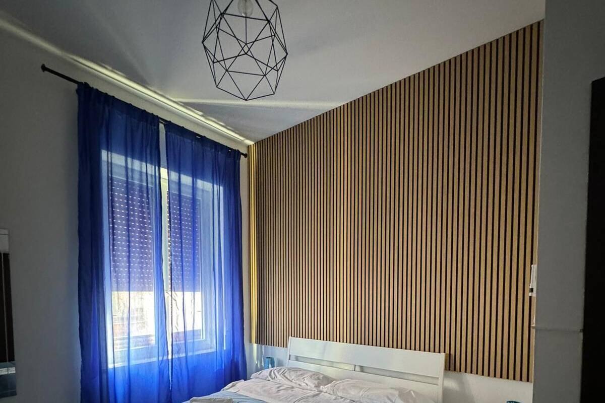Quarto moderno com cama, mesa de cabeceira, luminária pendente e janela com cortinas azuis.