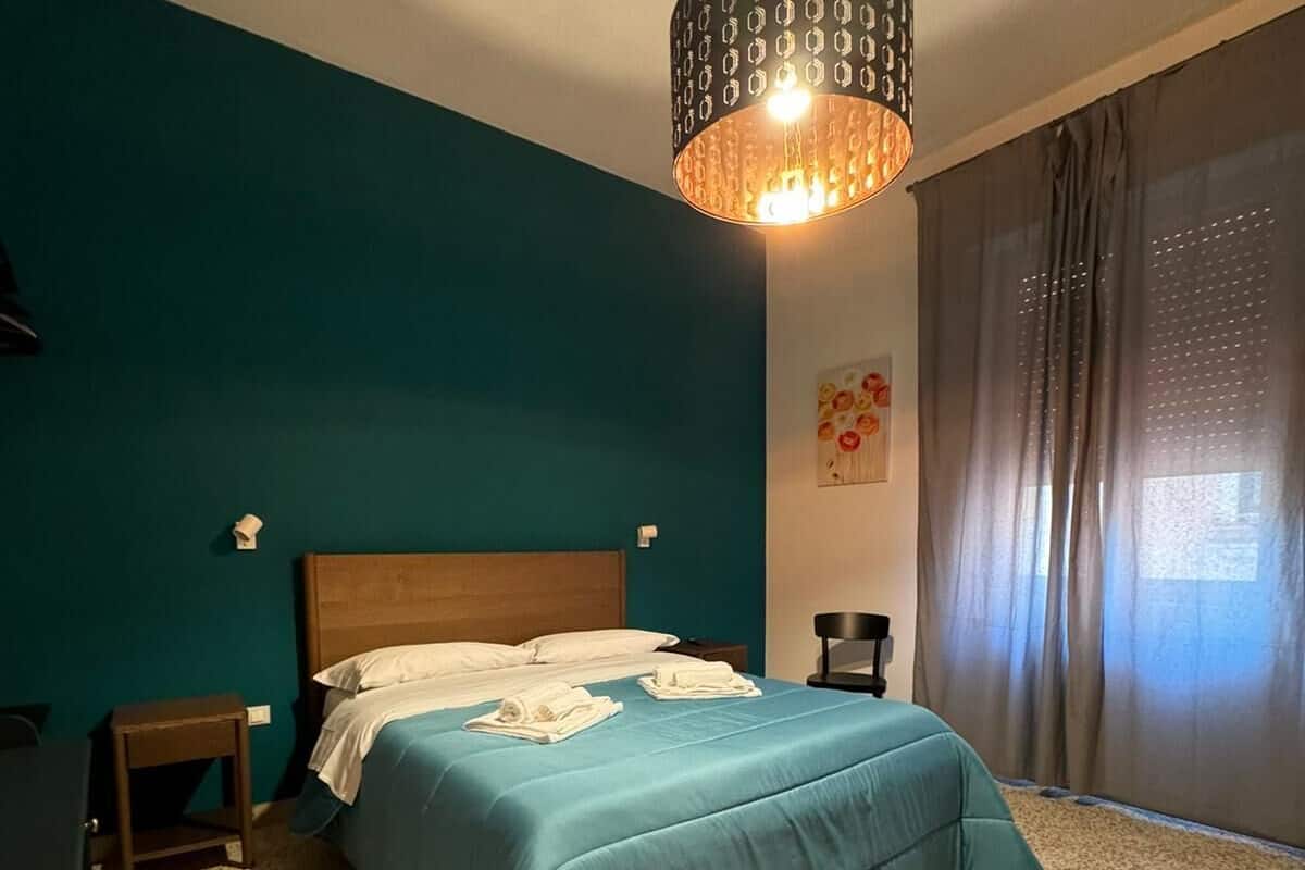 Quarto com parede verde-água, cabeceira de madeira, cama com roupa de cama azul, luminária de chão estampada e janela com cortinas.